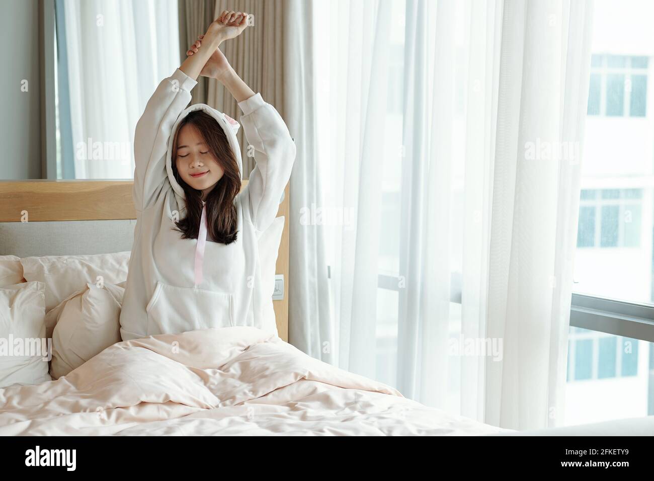 Beautiful smiling young Asian woman stretching in bed after waking up ...