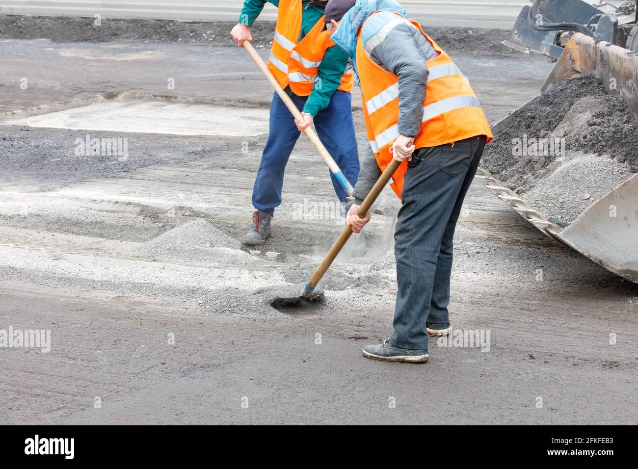 Two road workers rake old broken asphalt off the road and load it into ...