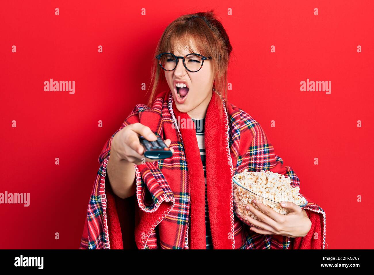 Redhead young woman eating popcorn using tv control angry and mad ...