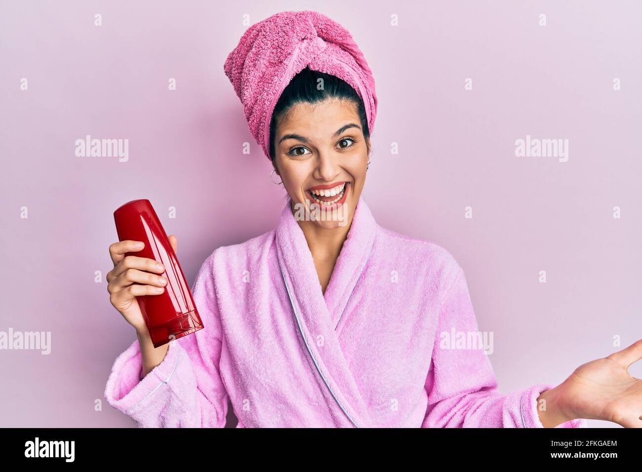 Young hispanic woman wearing shower bathrobe holding shampoo ...