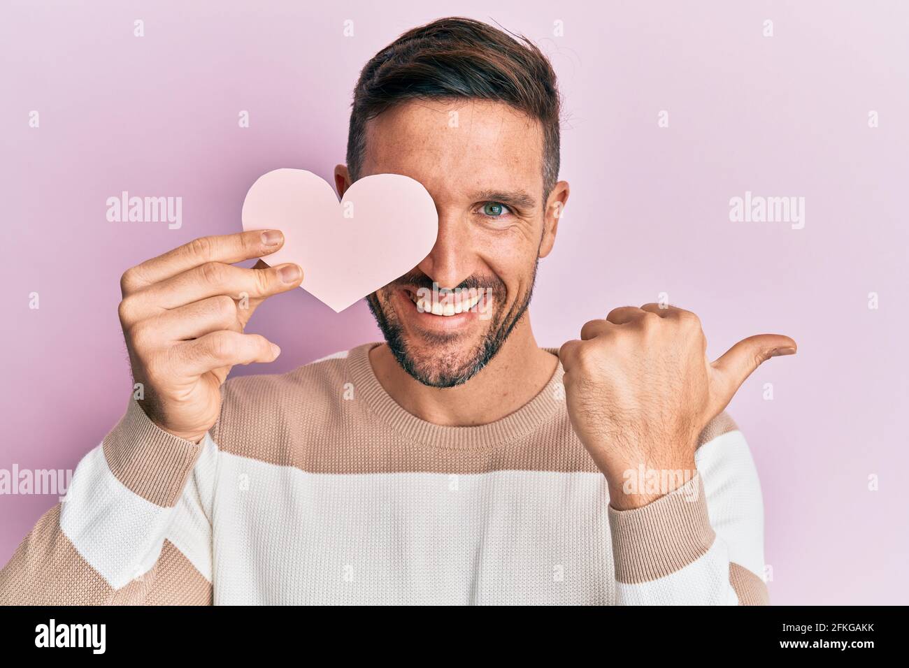 Handsome man with beard holding heart shape paper pointing thumb up to ...