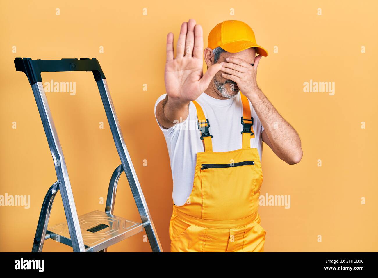 Handsome middle age man with grey hair holding ladder covering eyes ...