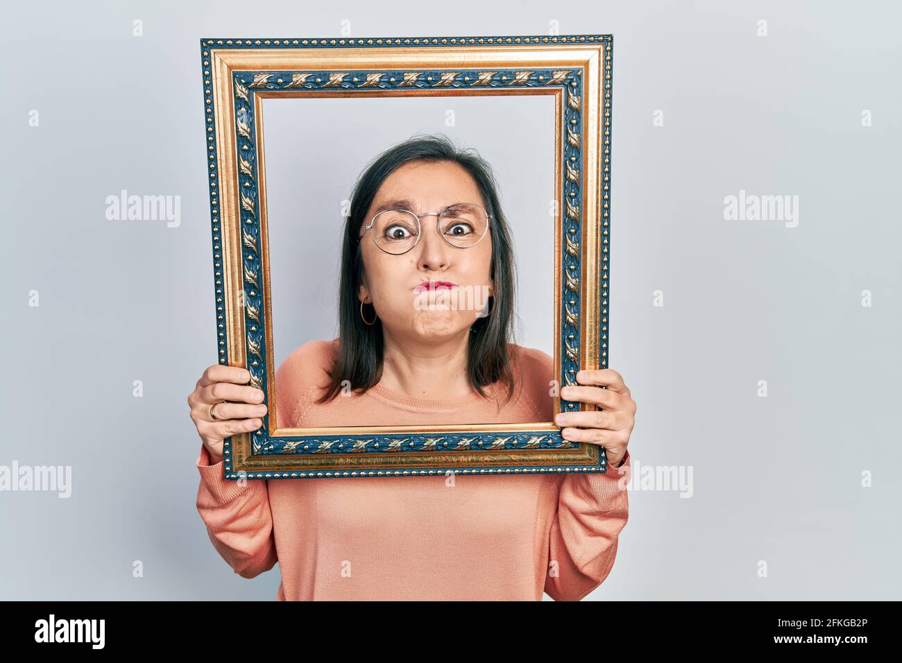 Middle age hispanic woman holding empty frame puffing cheeks with funny ...