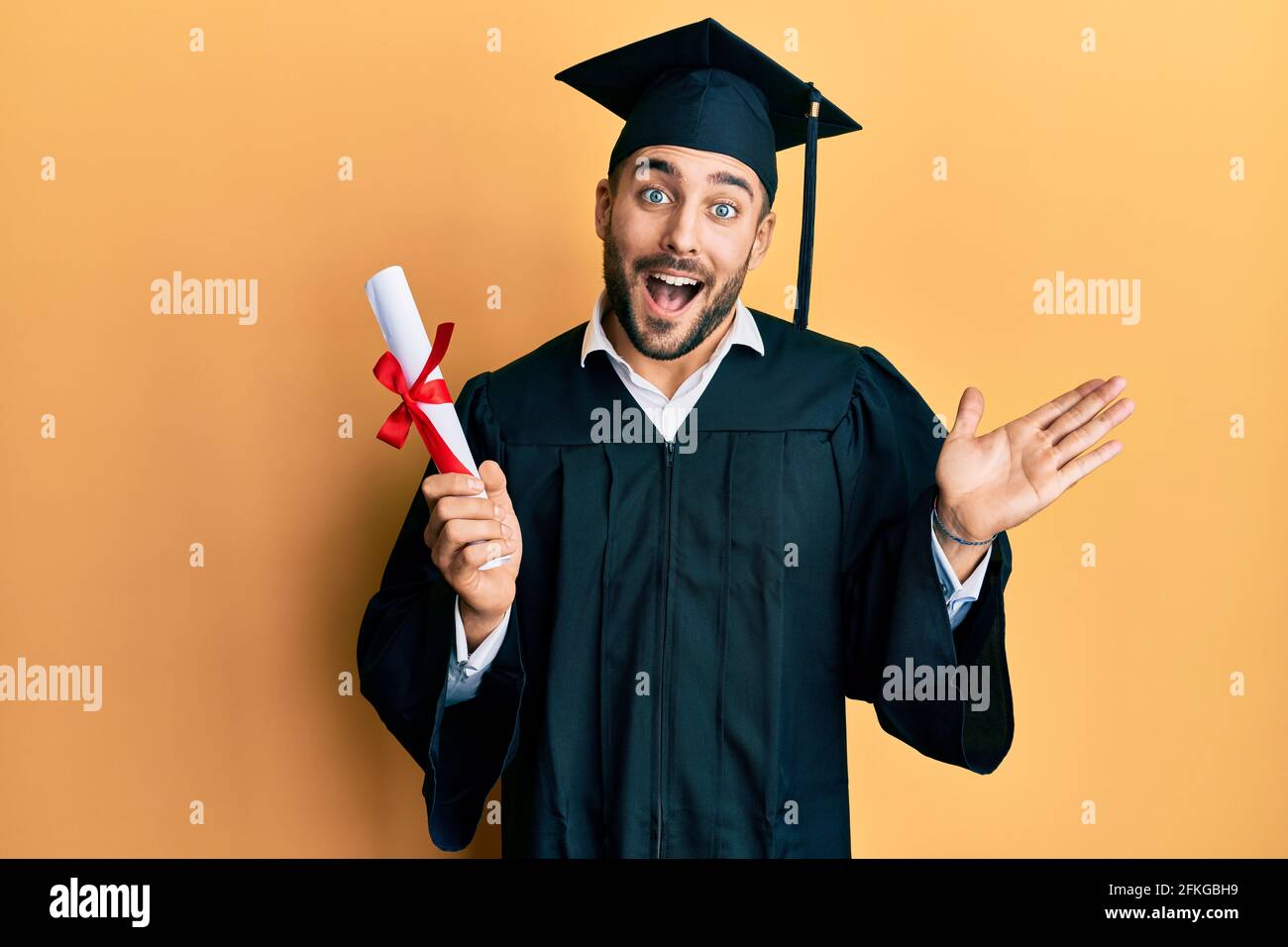 Young hispanic man wearing graduation robe holding diploma celebrating ...