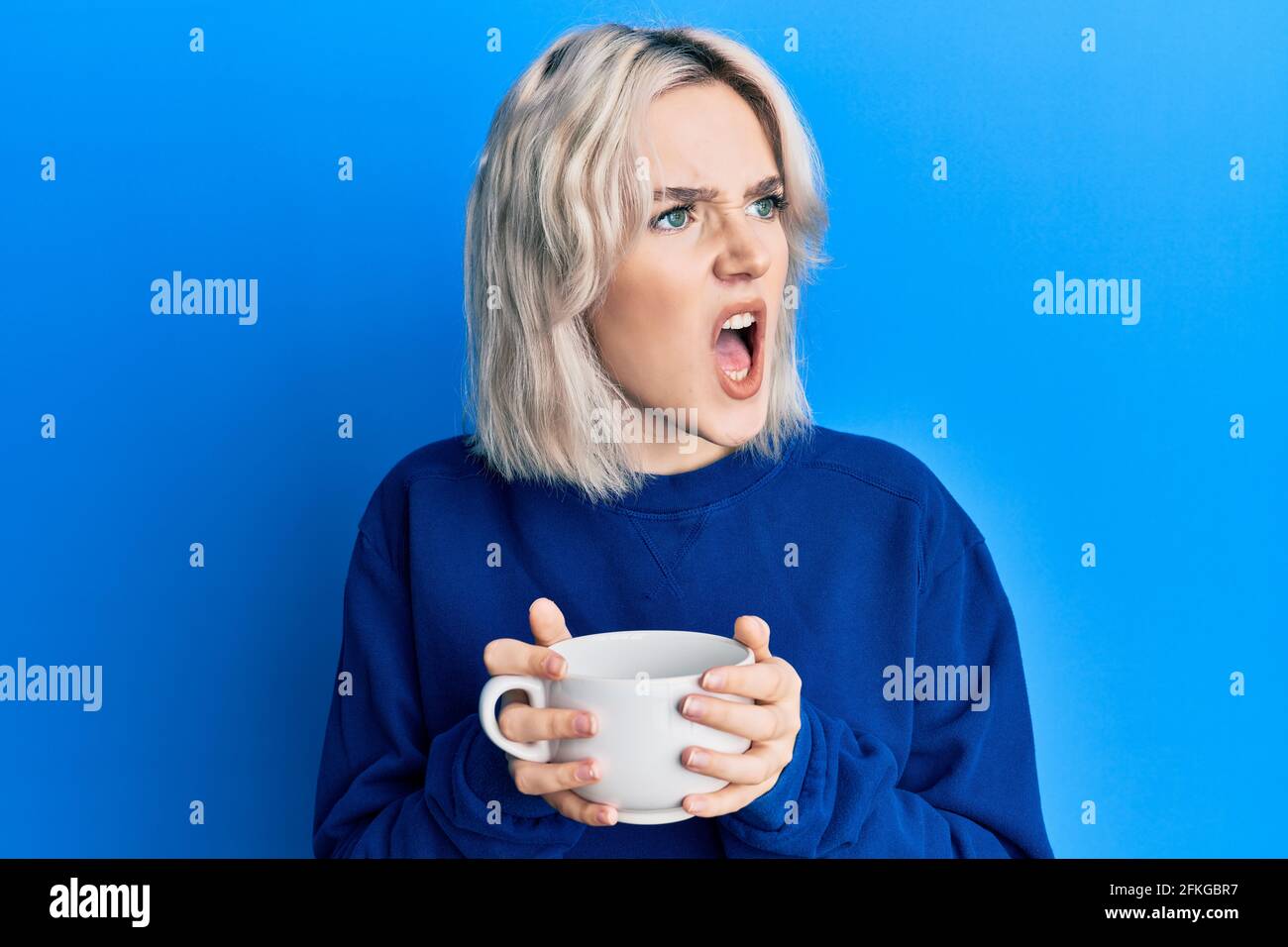 Young blonde girl drinking a cup of coffee angry and mad screaming ...