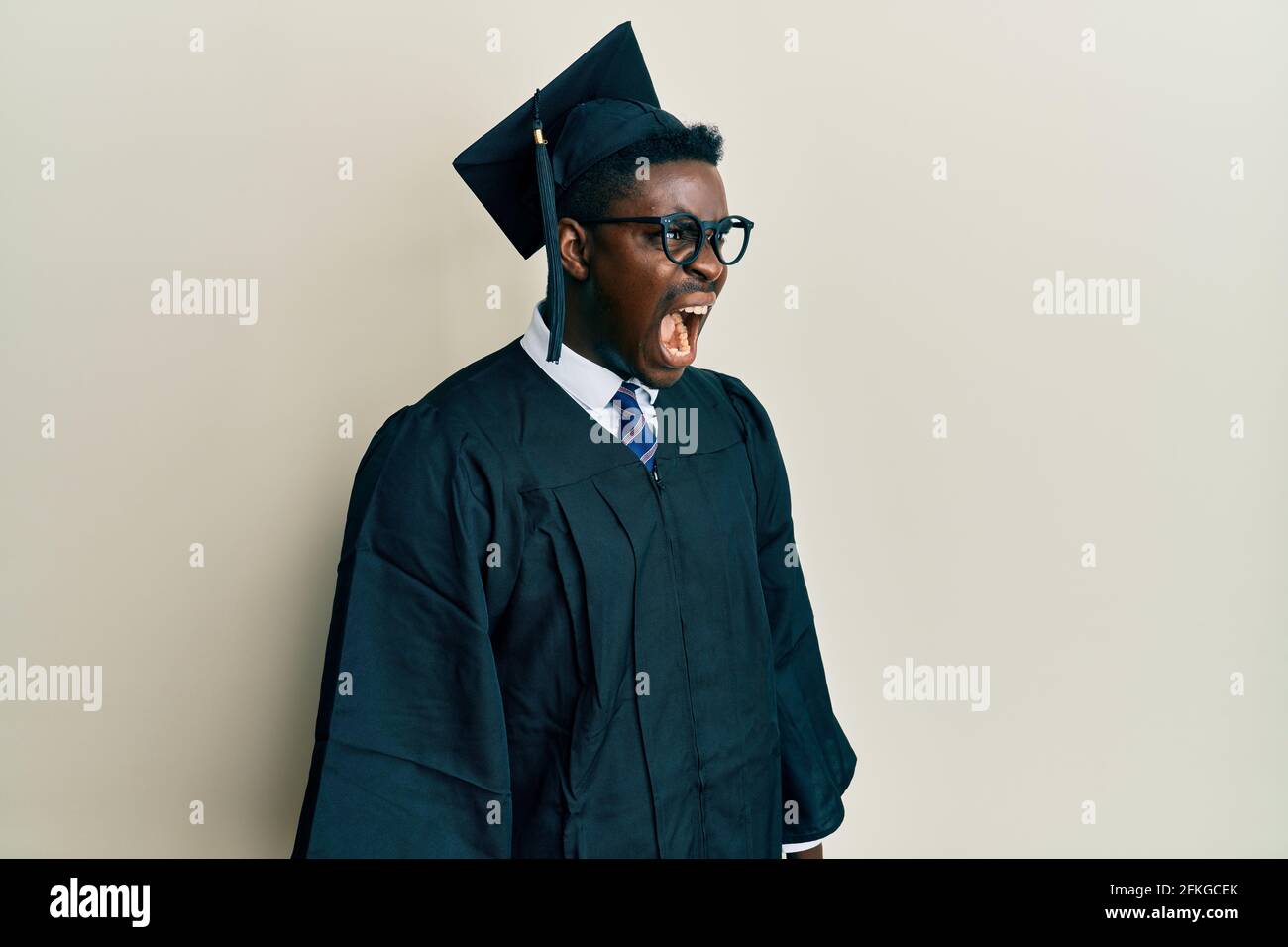 Handsome black man wearing graduation cap and ceremony robe angry and ...