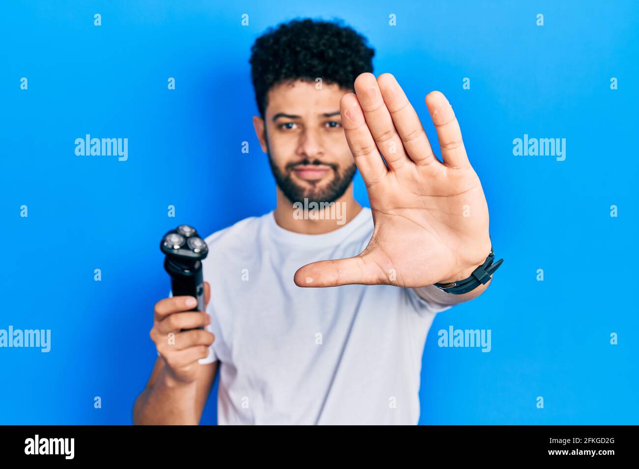 Young arab man with beard holding electric razor machine with open hand ...