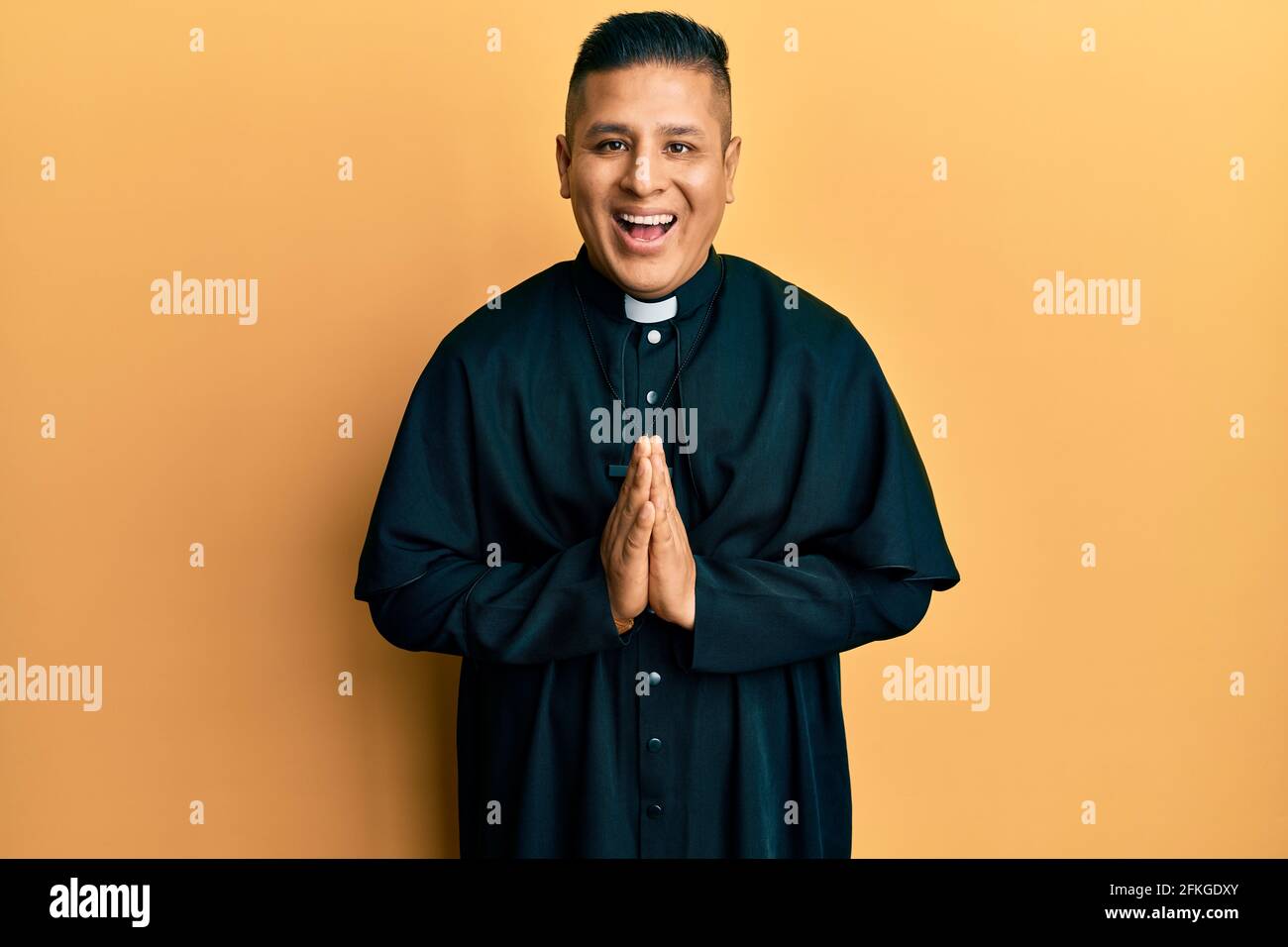Young latin priest man praying with hands together smiling and laughing ...