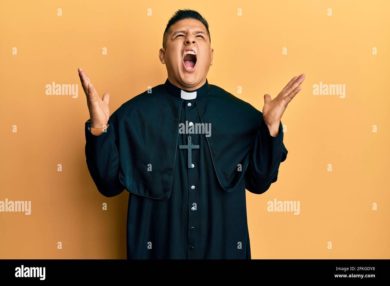 Young latin priest man standing over yellow background crazy and mad ...