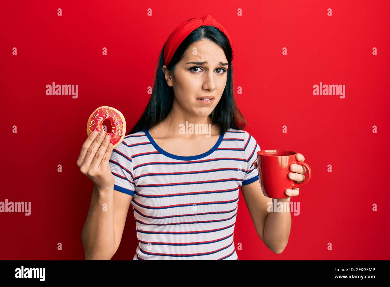 Young hispanic woman eating doughnut and drinking coffee clueless and ...
