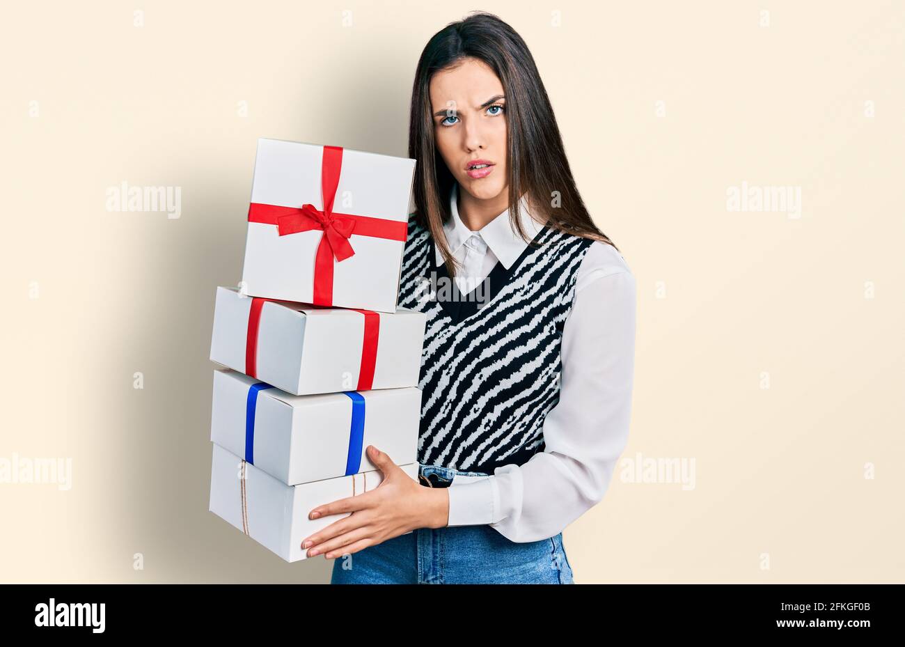 Young brunette teenager holding gifts in shock face, looking skeptical ...
