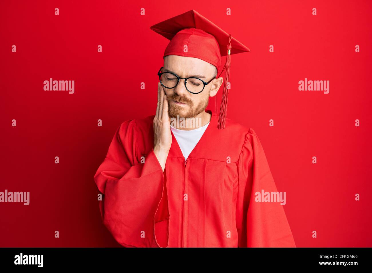 Young redhead man wearing red graduation cap and ceremony robe touching ...