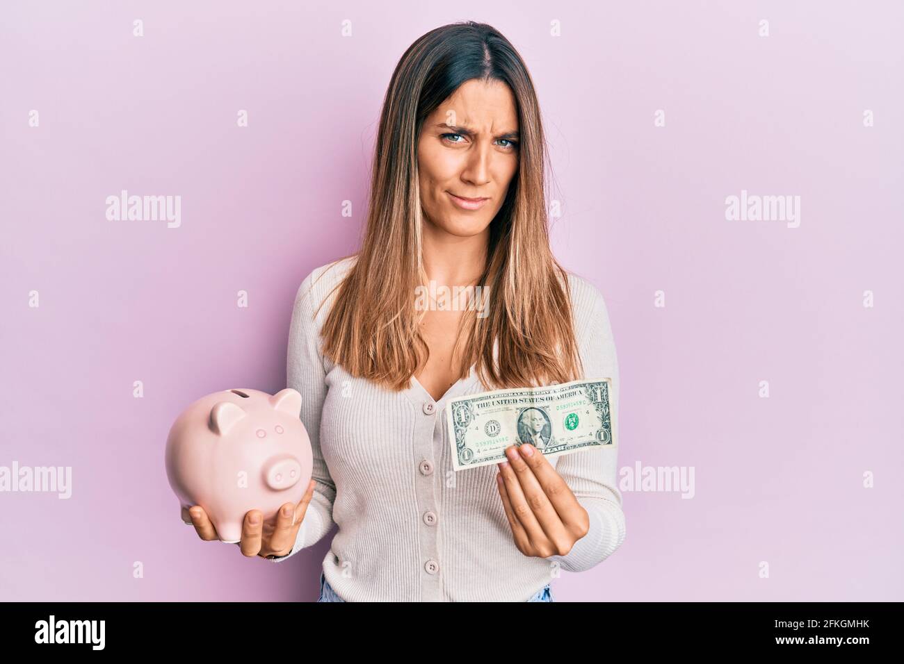 Brunette young woman holding one dollar banknote and piggy bank skeptic ...