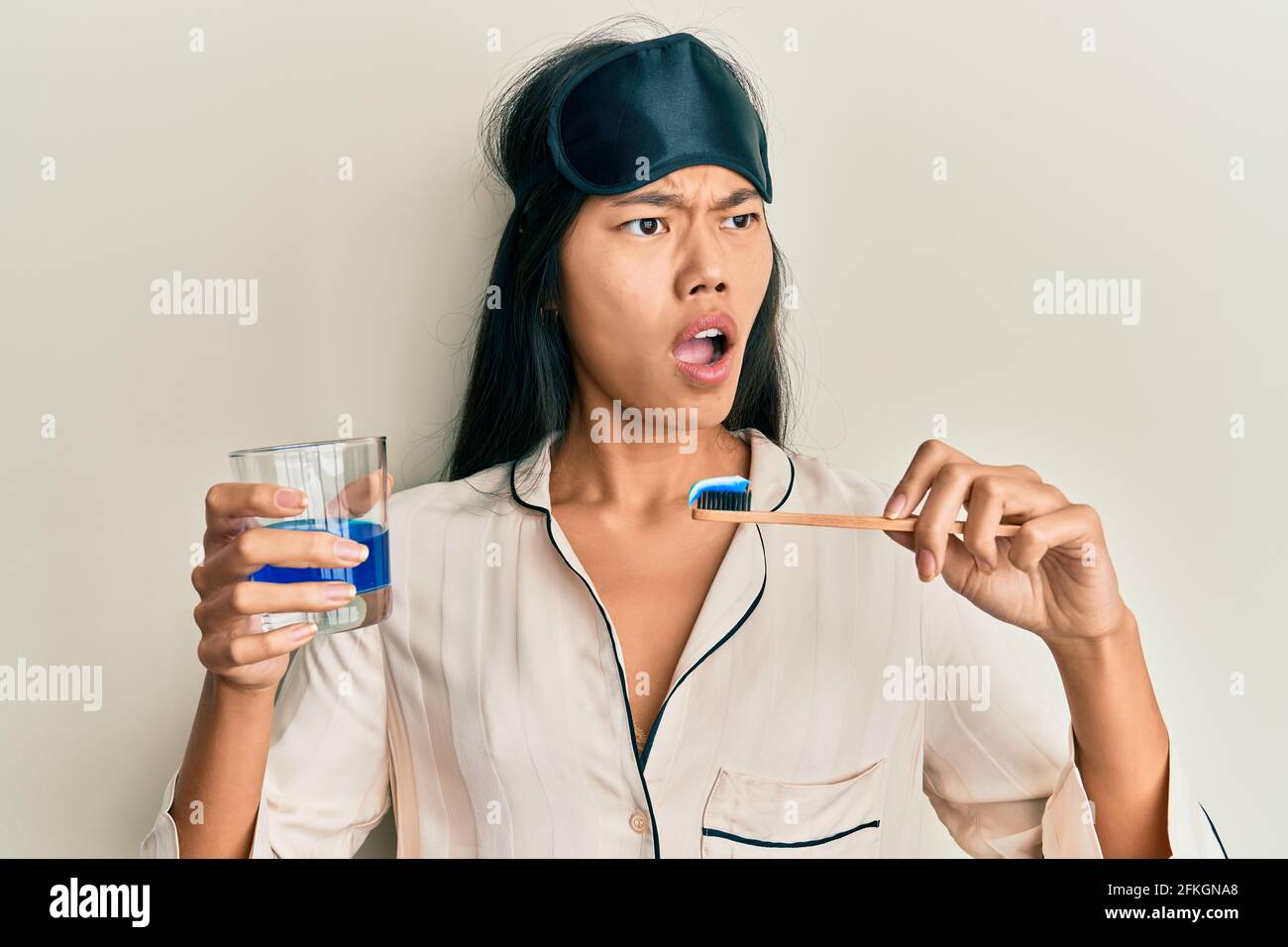 Young chinese woman wearing pajama using toothbrush and mouthwash angry ...