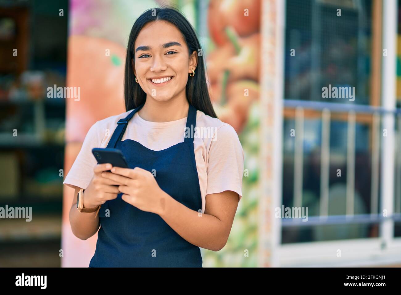 Young latin shopkeeper girl smiling happy using smartphone at fruit ...