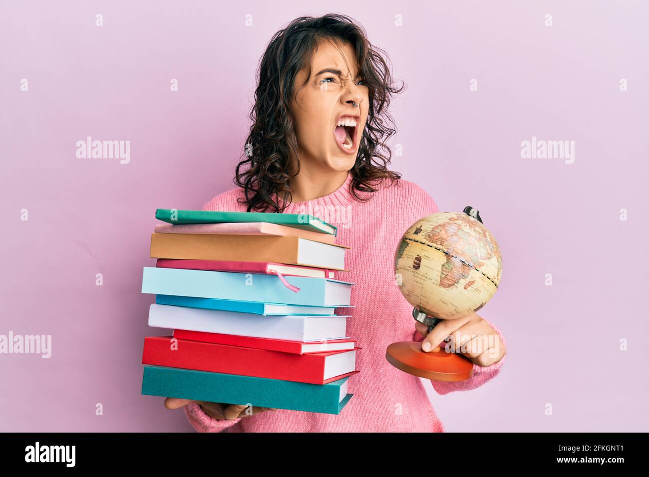 Young hispanic woman holding a pile of books and world ball angry and ...