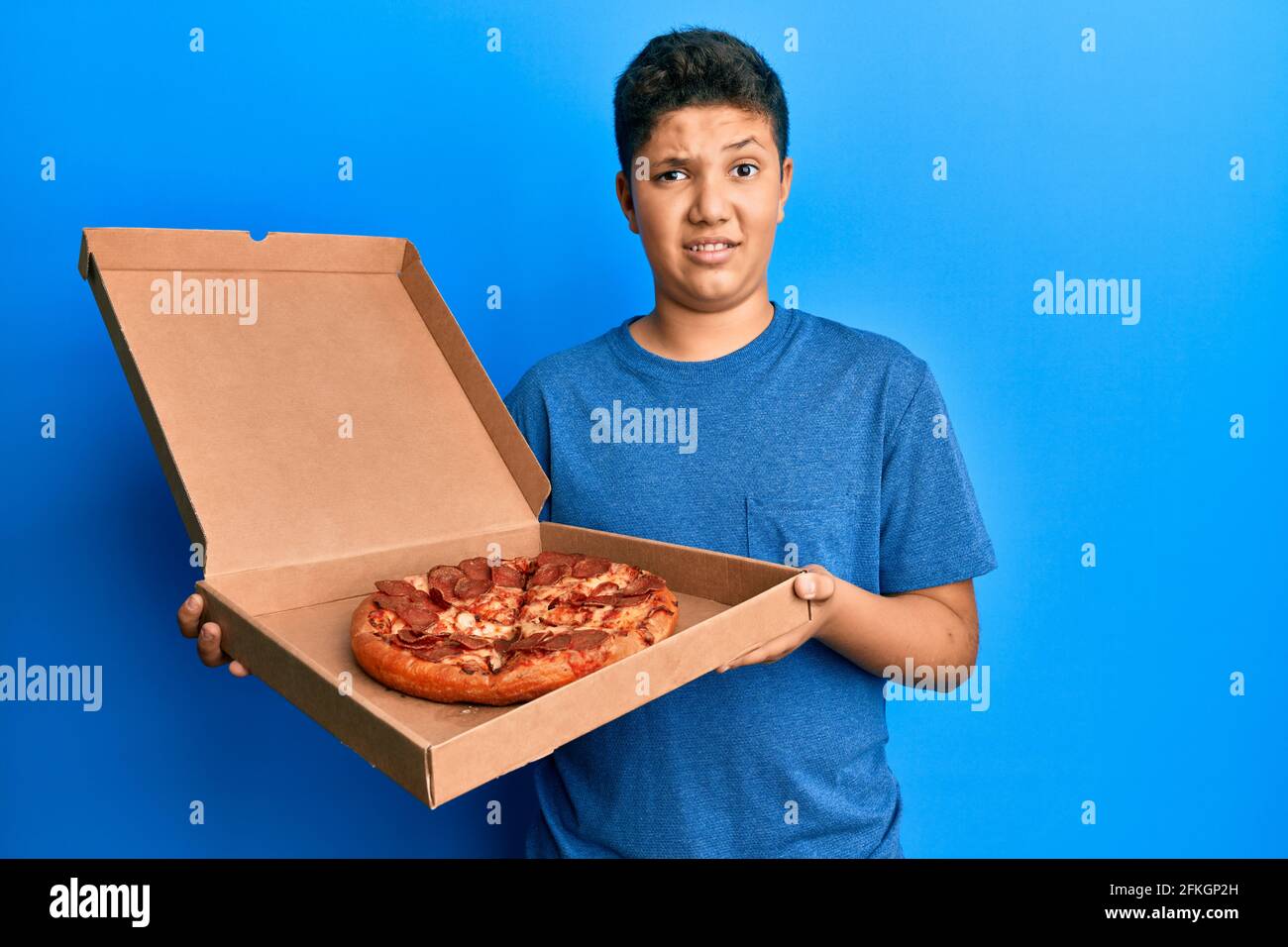 Teenager hispanic boy eating tasty pepperoni pizza skeptic and nervous ...