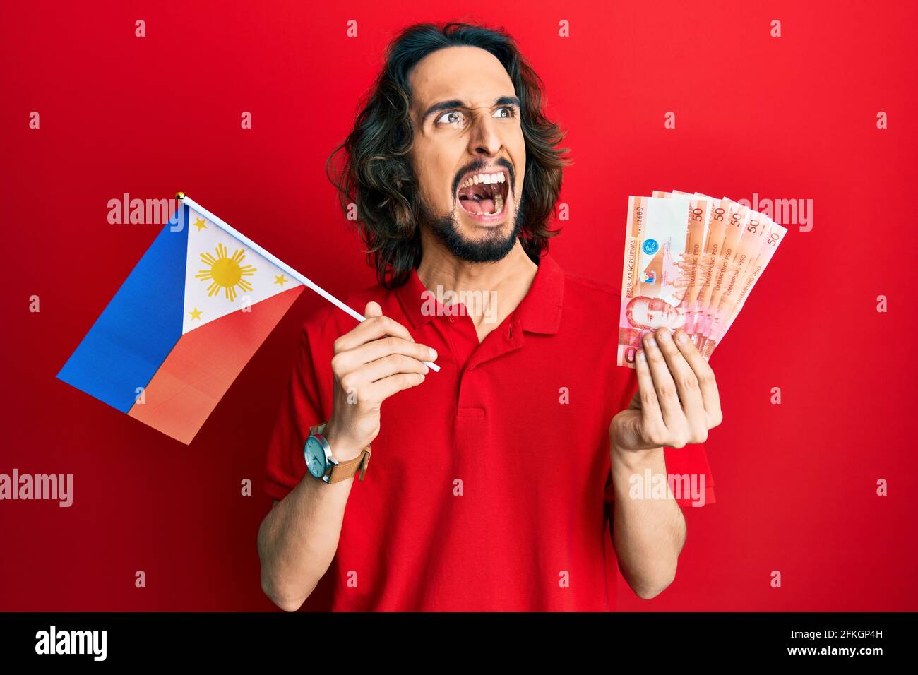 Young hispanic man holding philippines flag and pesos banknotes angry ...