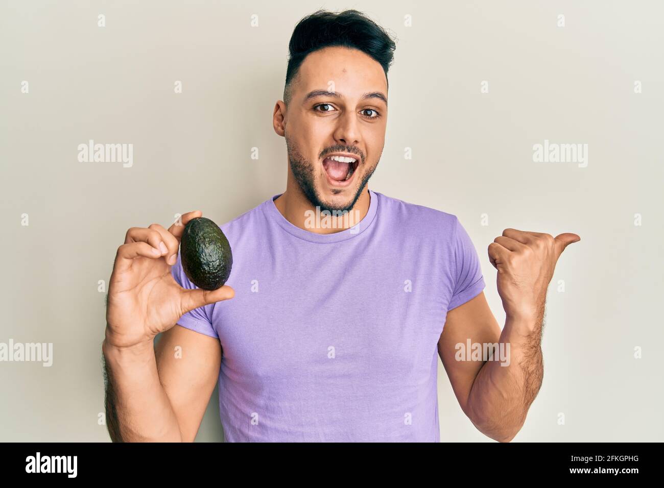 Young arab man holding avocado pointing thumb up to the side smiling ...