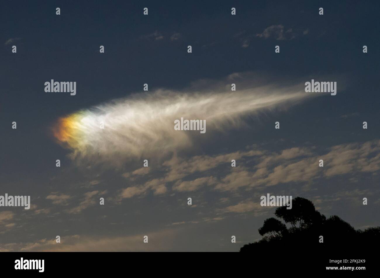 Small white cloud with rainbow of refracted light at one end. In dark ...