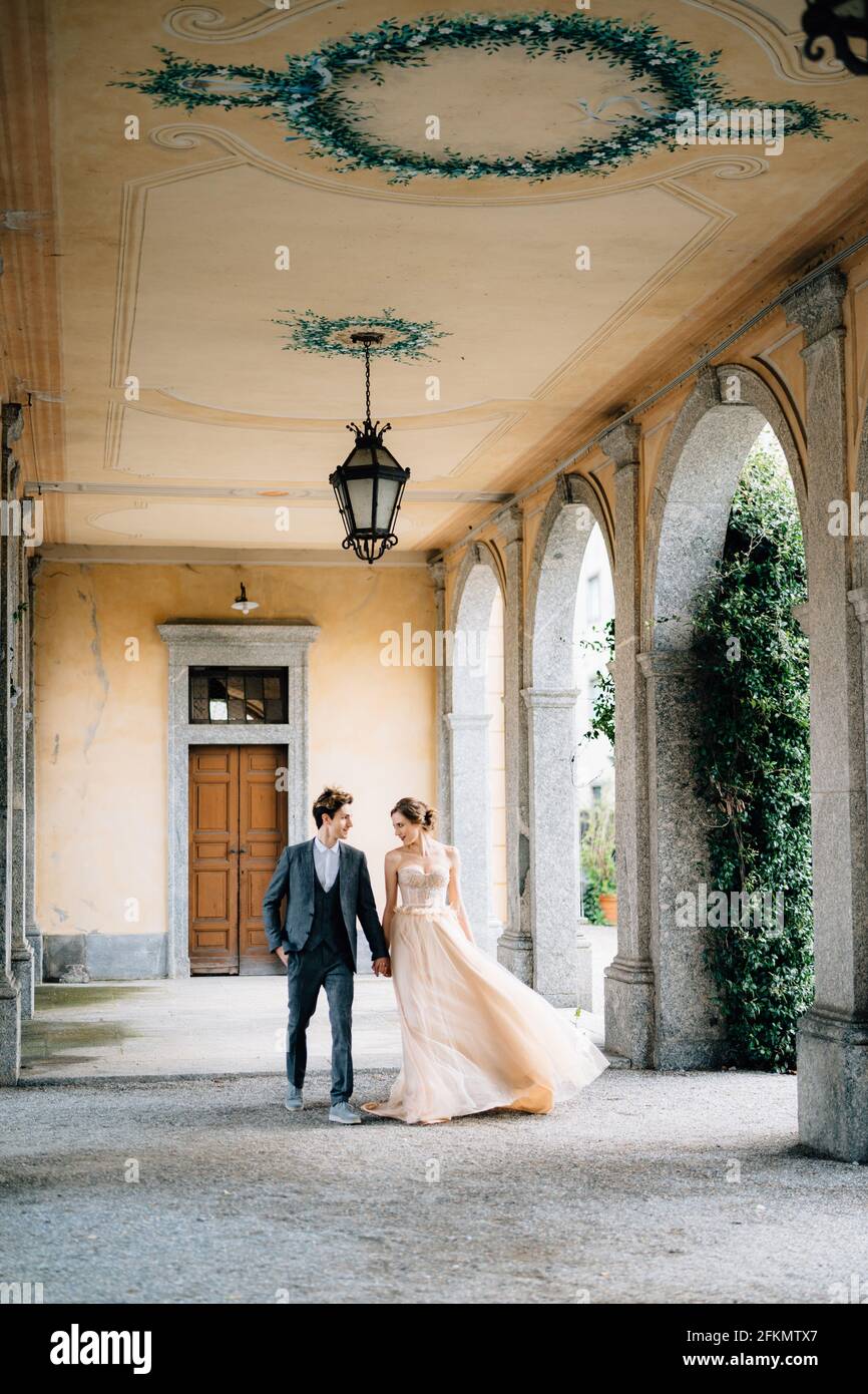 Bride and groom walk along the old terrace with columns entwined with ...