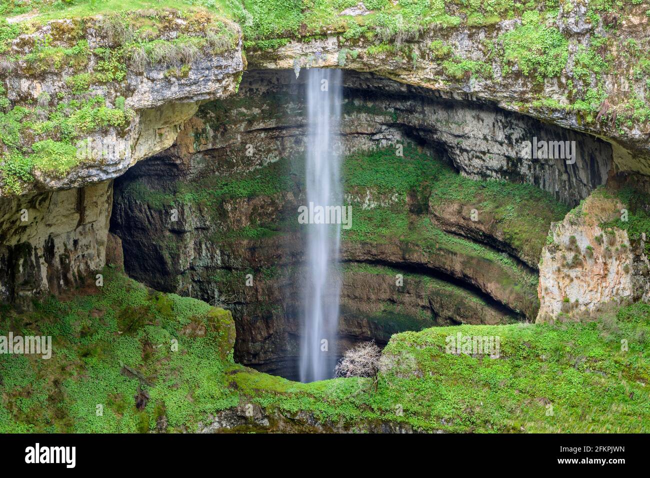 Waterfall behind a natural bridge covered in lush green vegetation ...