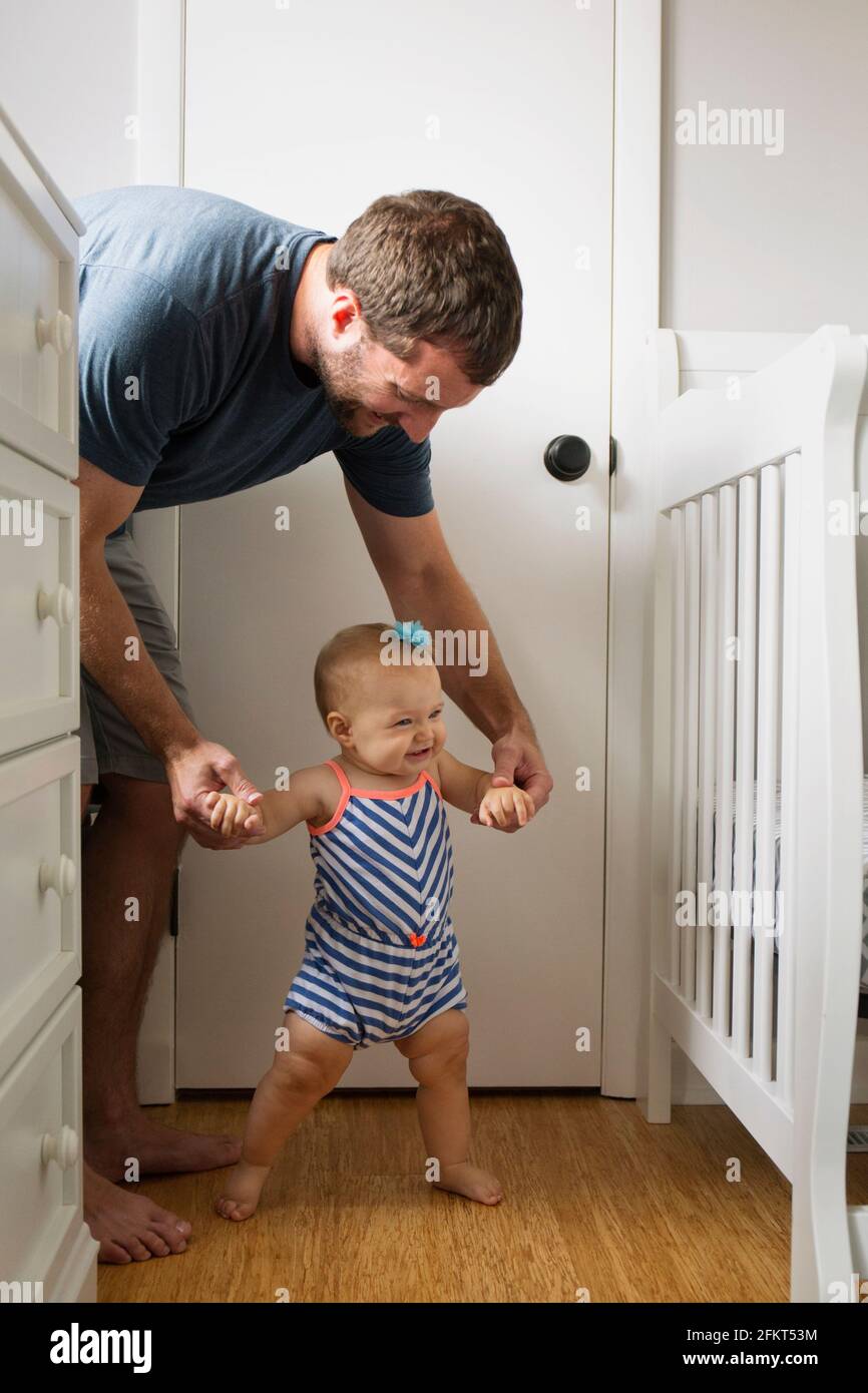 Mid adult man holding baby daughter hands whilst toddling in nursery ...
