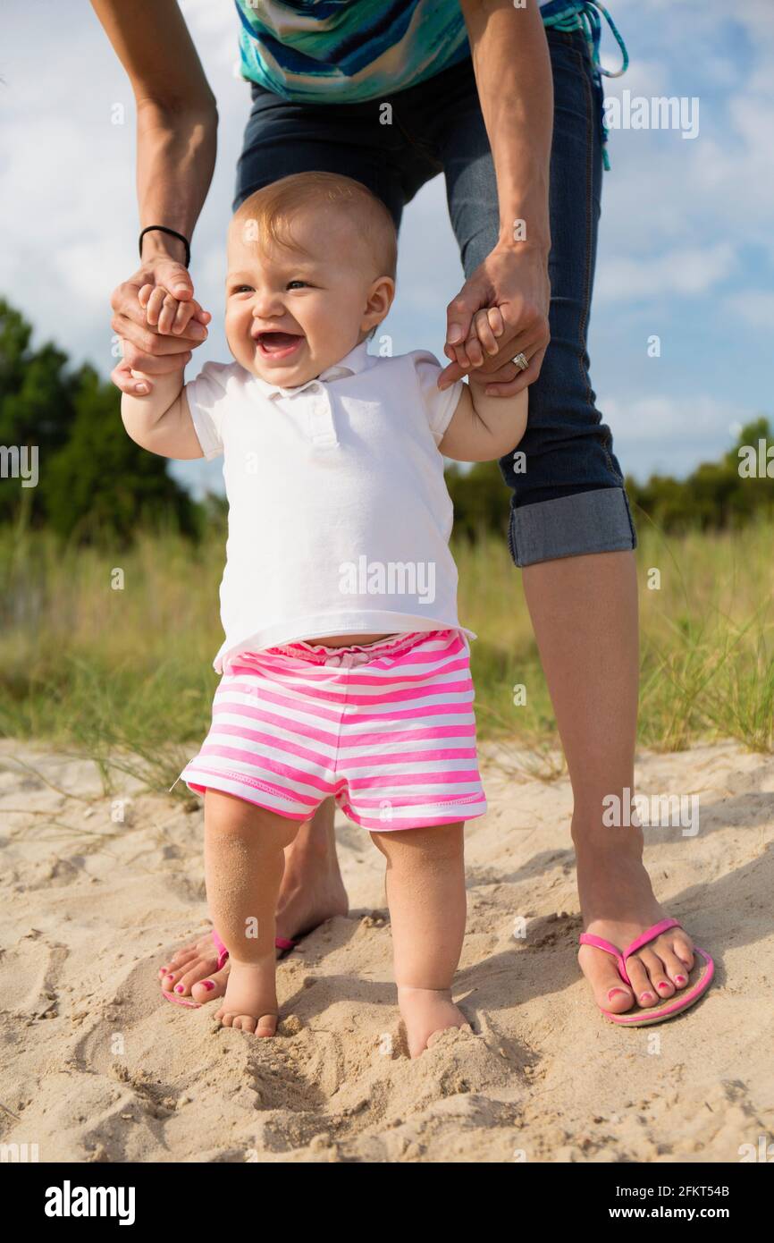 Mid adult woman holding baby daughters hands while toddling in sand ...