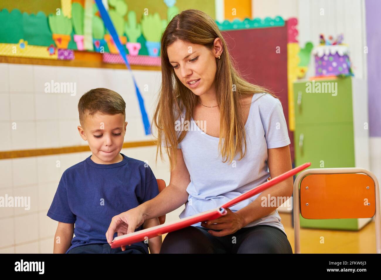Educator or childminder with child reading a book in kindergarten or ...
