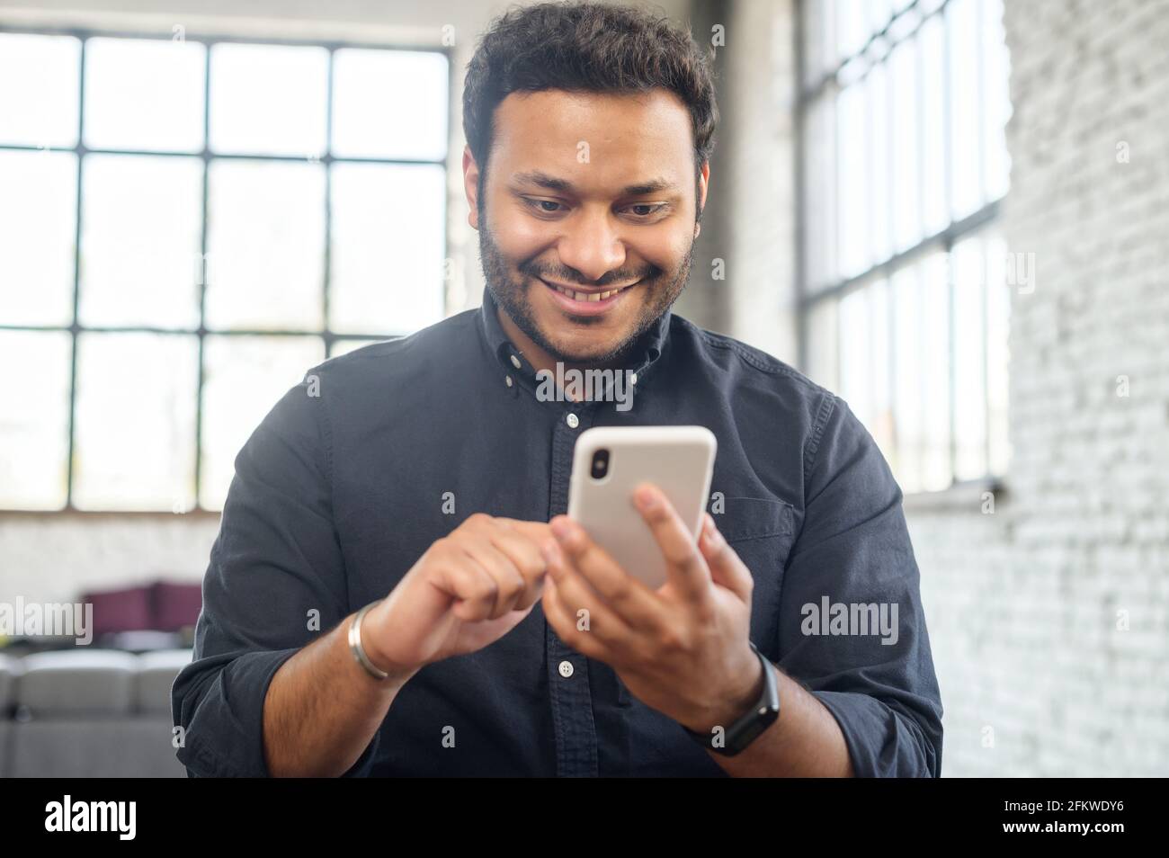 Positive indian guy using smartphone for online communication, smiling mixed-race guy browsing ...