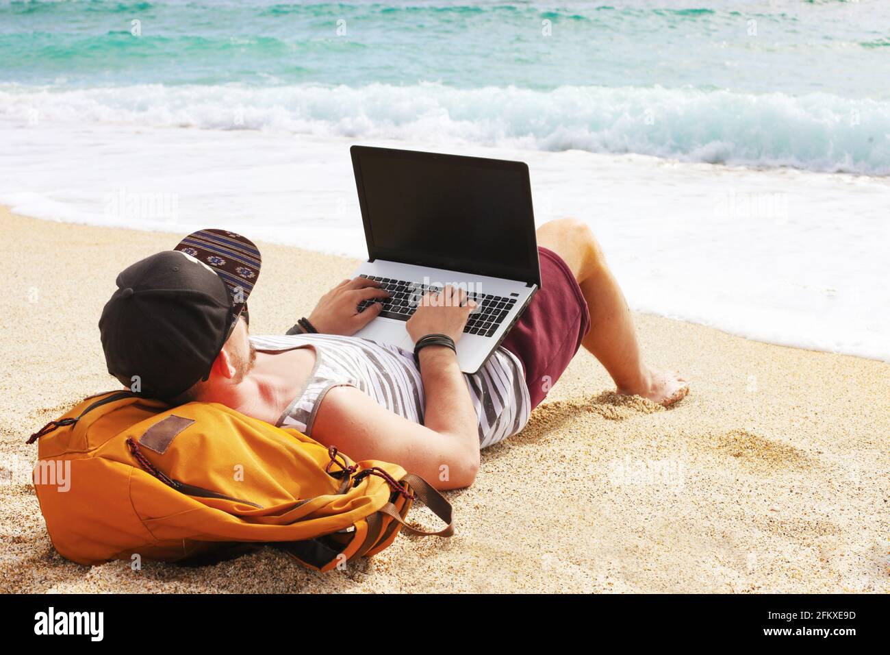 Back view, hipster young man lying on sandy beach leaning on backpack ...