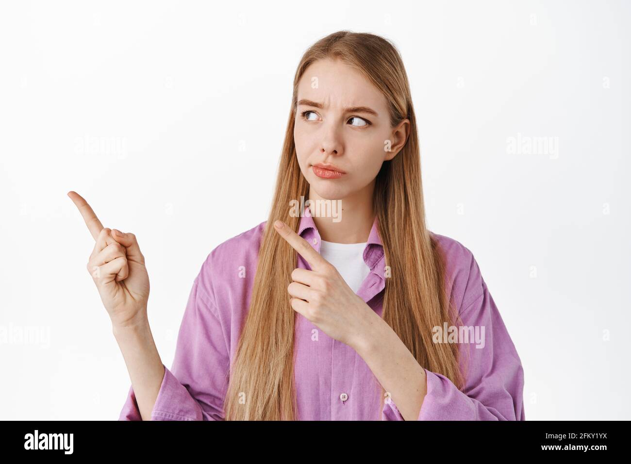 Thoughtful young woman looking suspicious aside, pointing fingers left ...