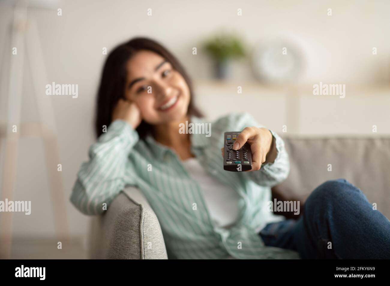 Happy Indian lady switching TV channels with remote control on sofa at ...