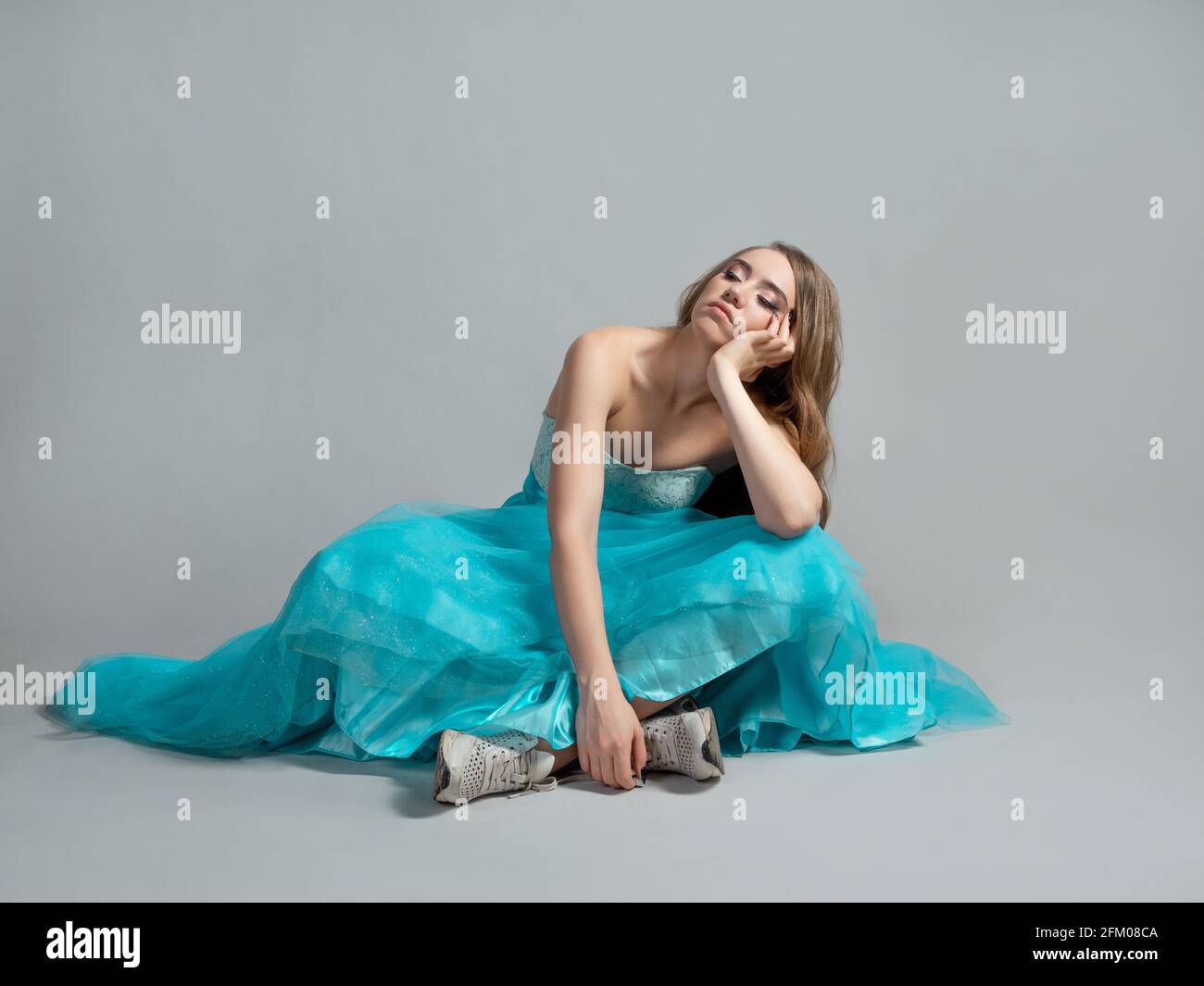 Bored princess in a lush blue dress sits on the floor, studio gray ...