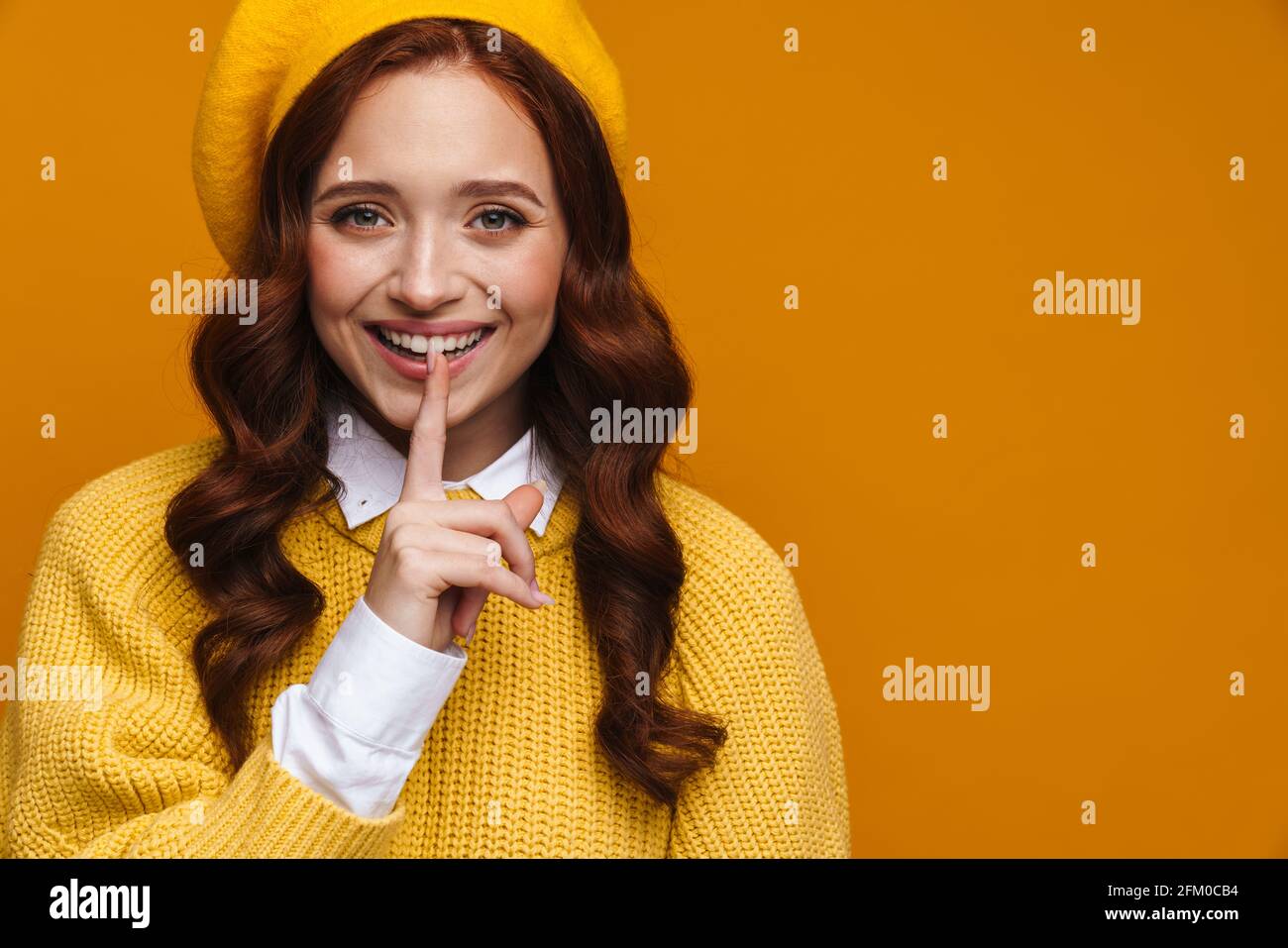 Happy young woman with long red hair in sweater and beret standing over yellow wall background ...