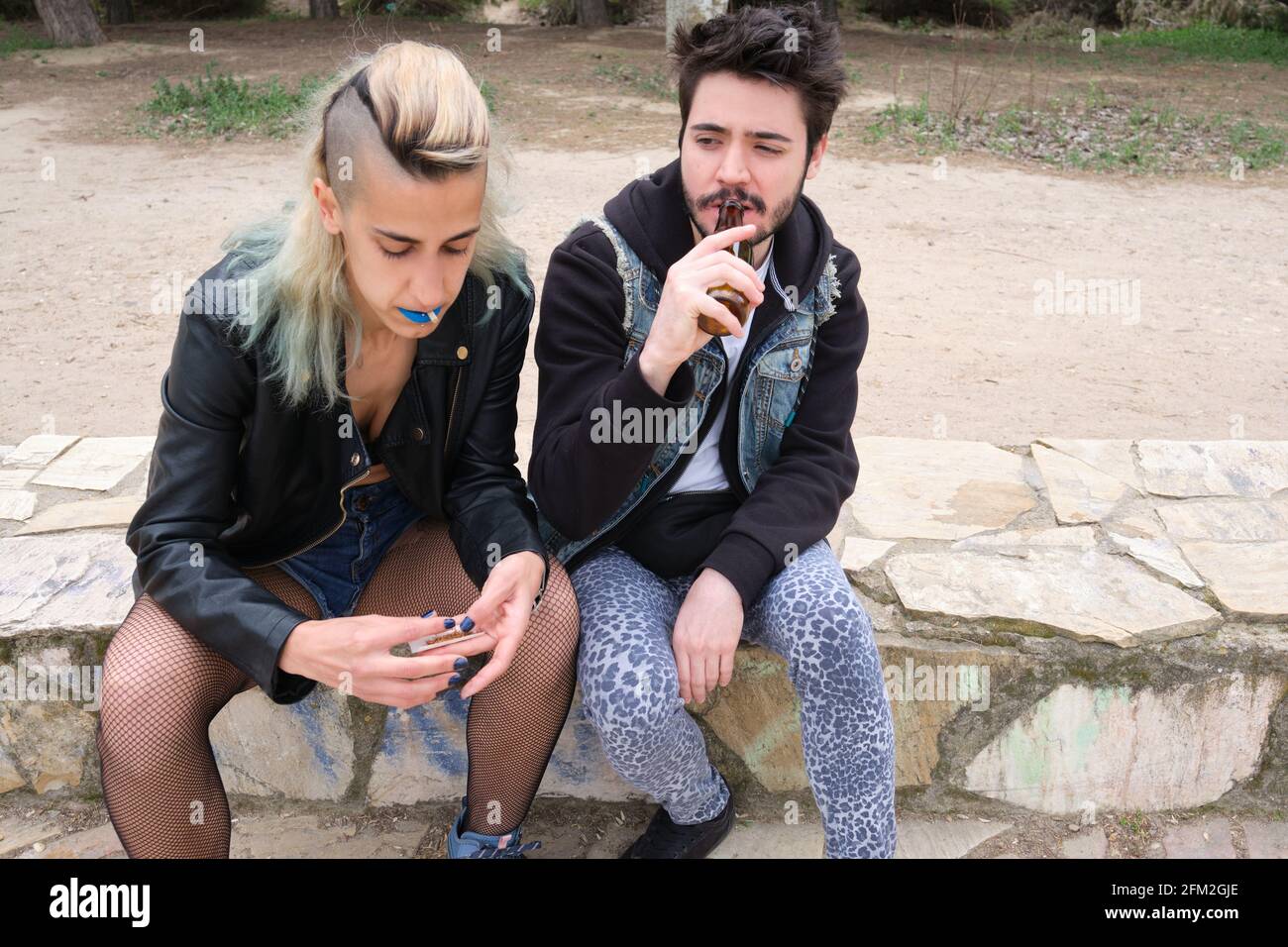 Young punk couple rolling a cigarette and drinking beer in a park. Rock ...