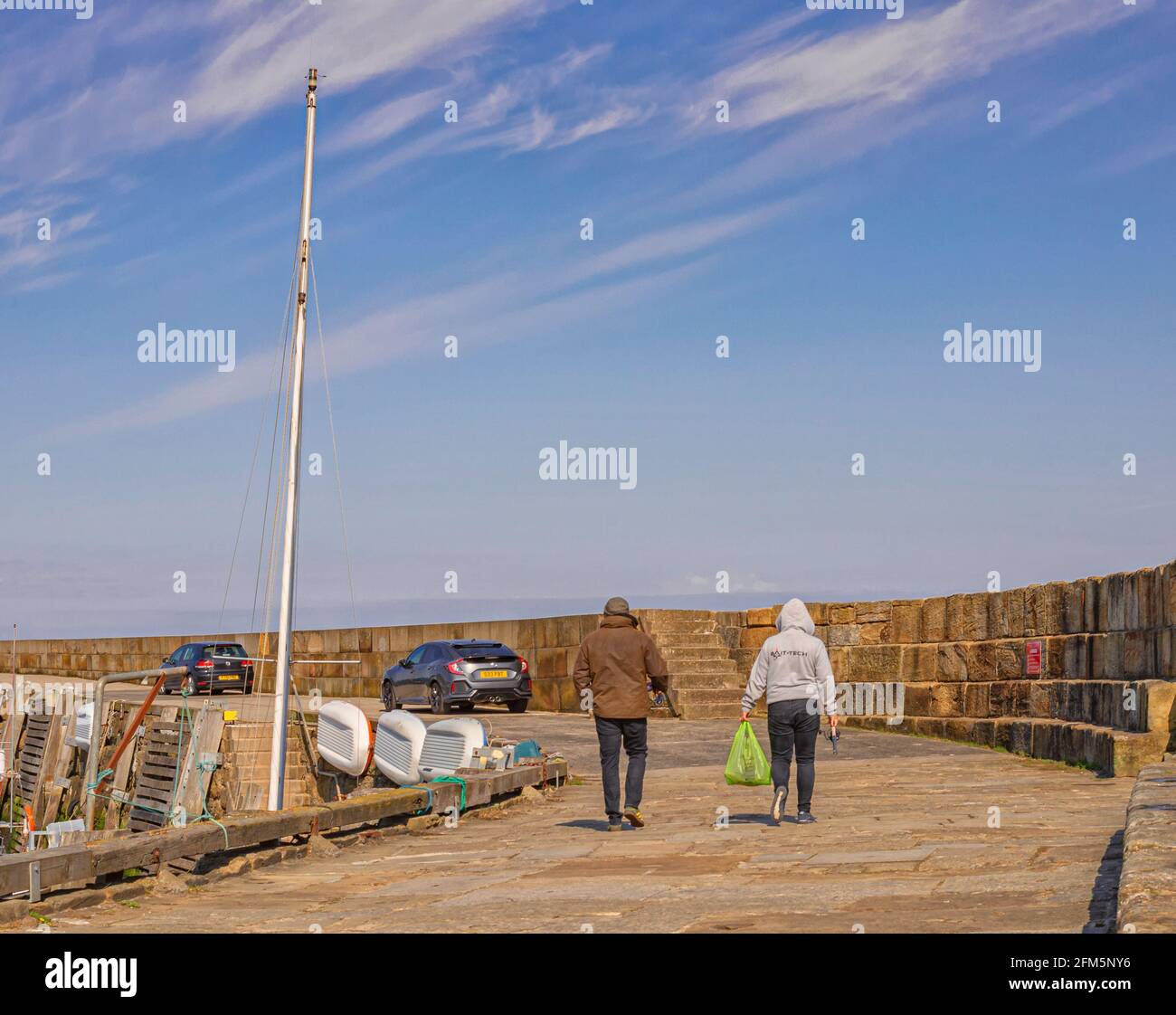 Two anglers walk along a harbour pier carrying their rods. A mast of a ...