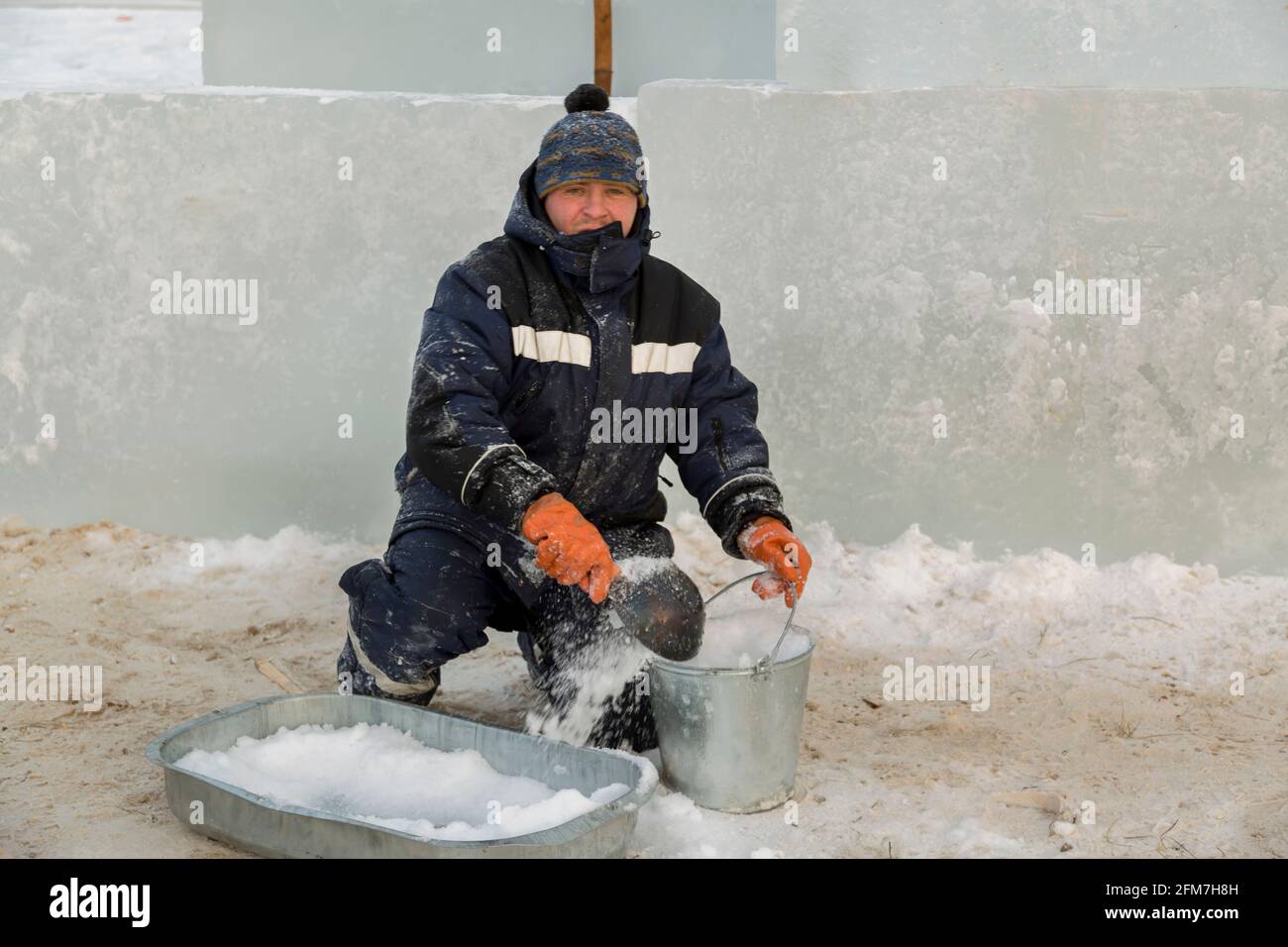 A worker assembler in winter overalls puts wet snow from a trough into ...