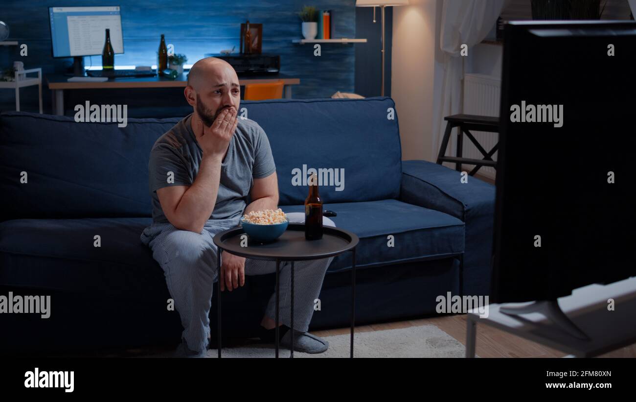 Focused man looking at drama movie, crying sitting on sofa eating ...