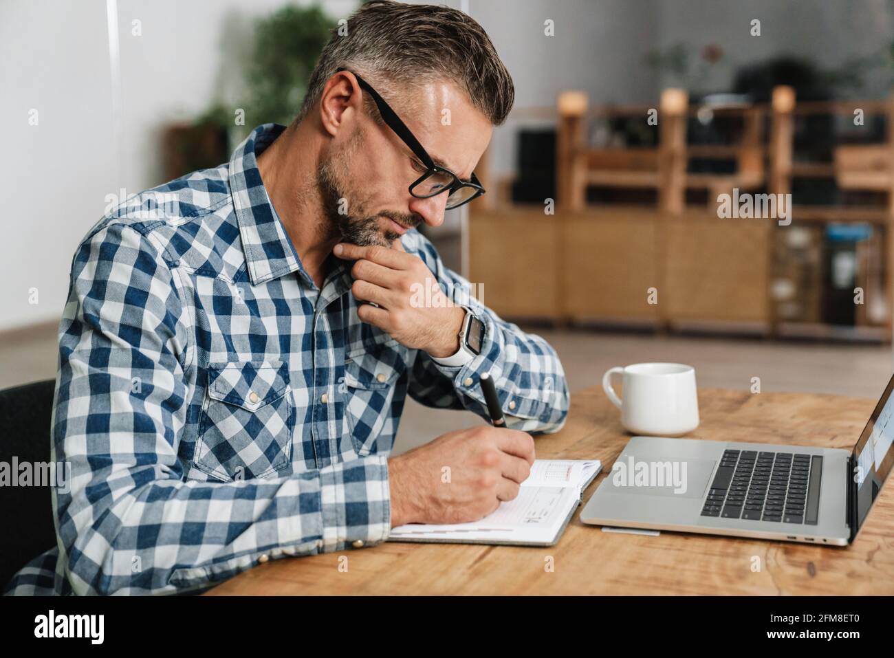 Focused grey man writing down notes while working with laptop in office ...