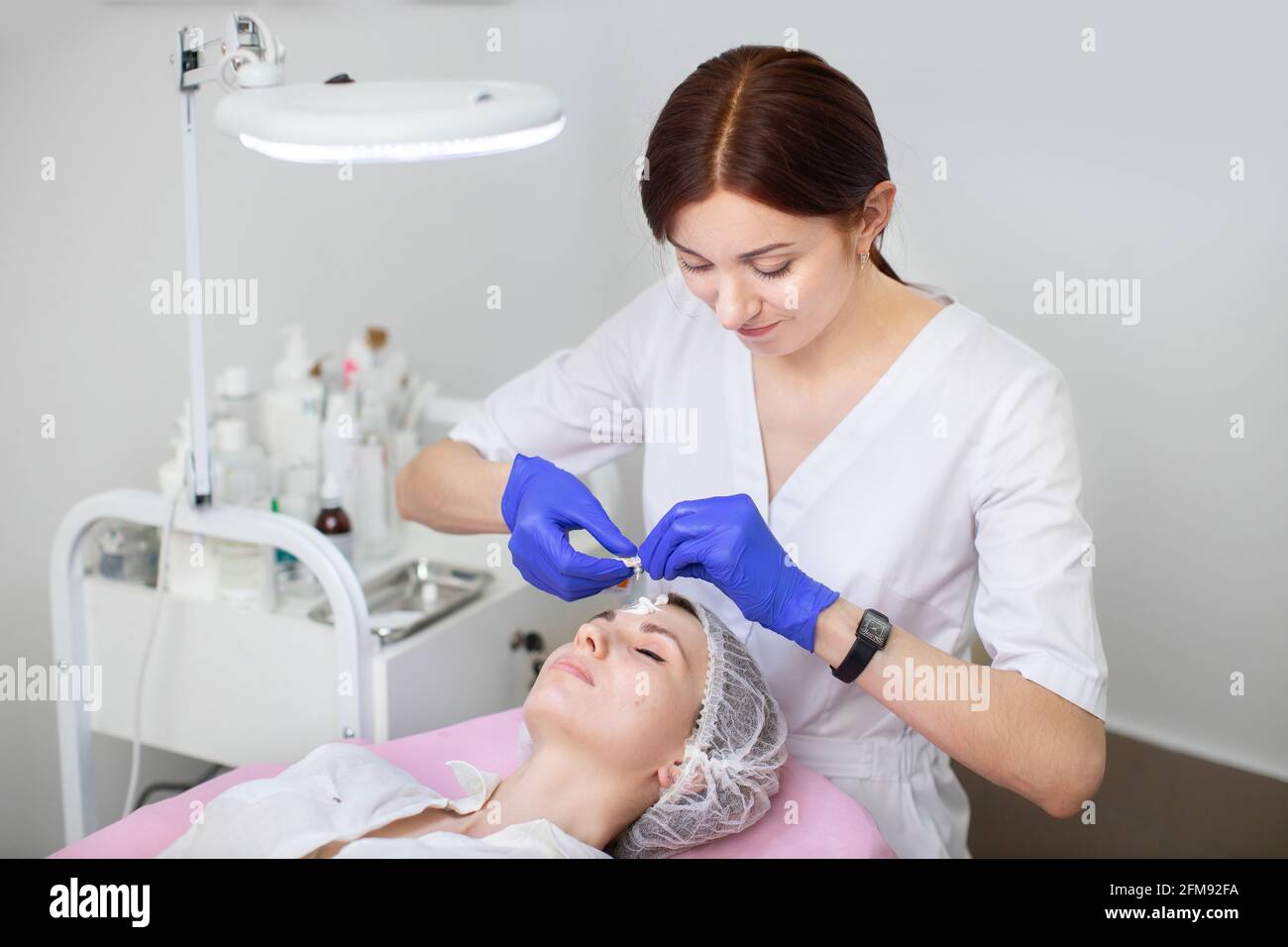 Female cosmetologist in a medical coat and rubber gloves applies a ...