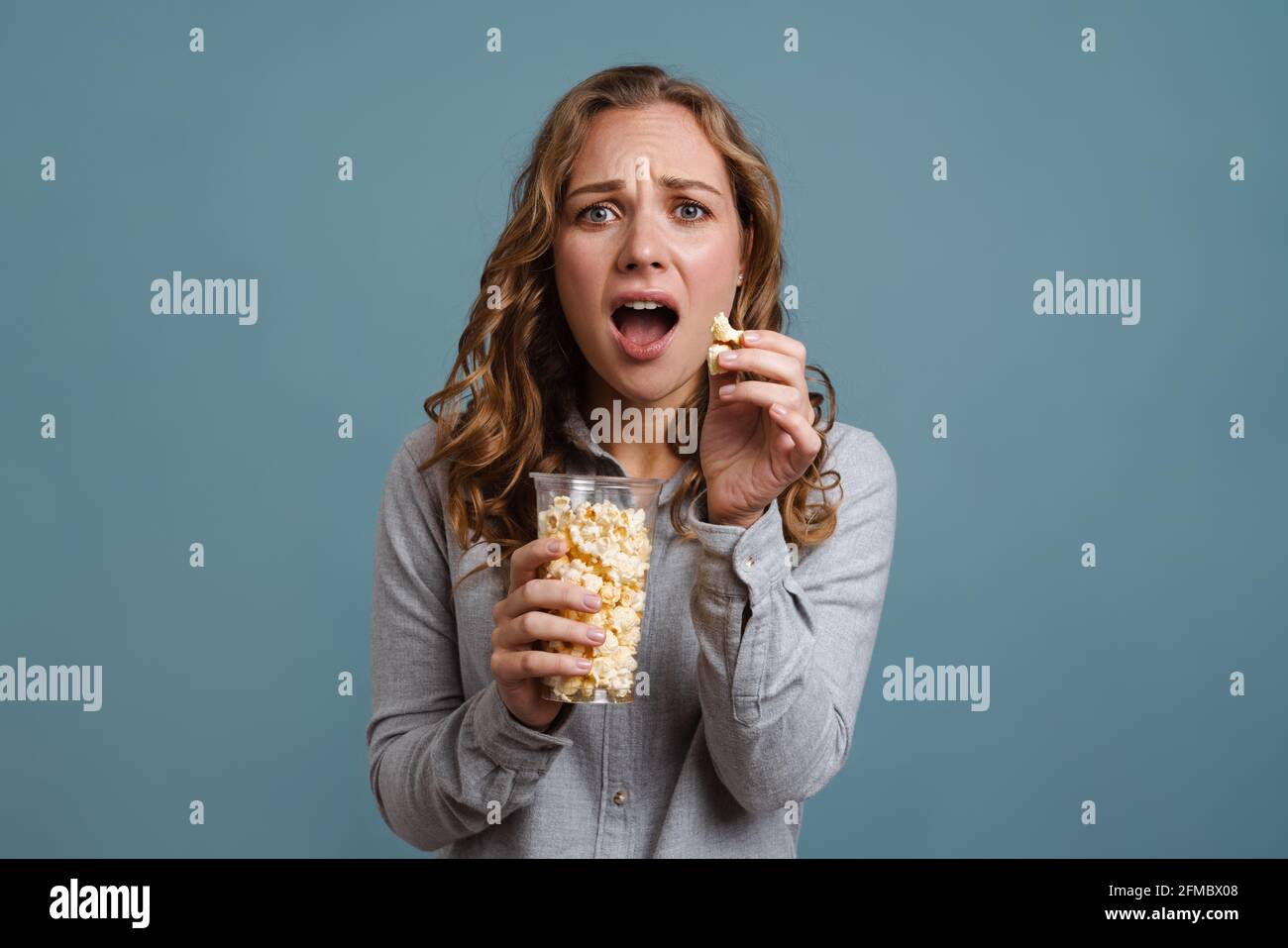 Shocked young woman watching TV and eating popcorn isolated over blue ...