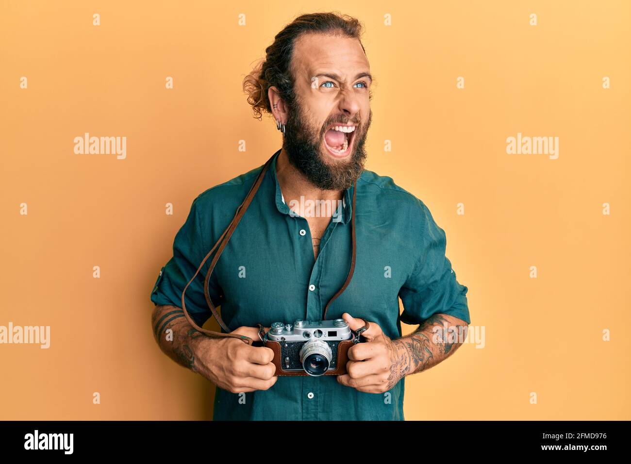 Handsome man with beard and long hair holding vintage camera angry and ...