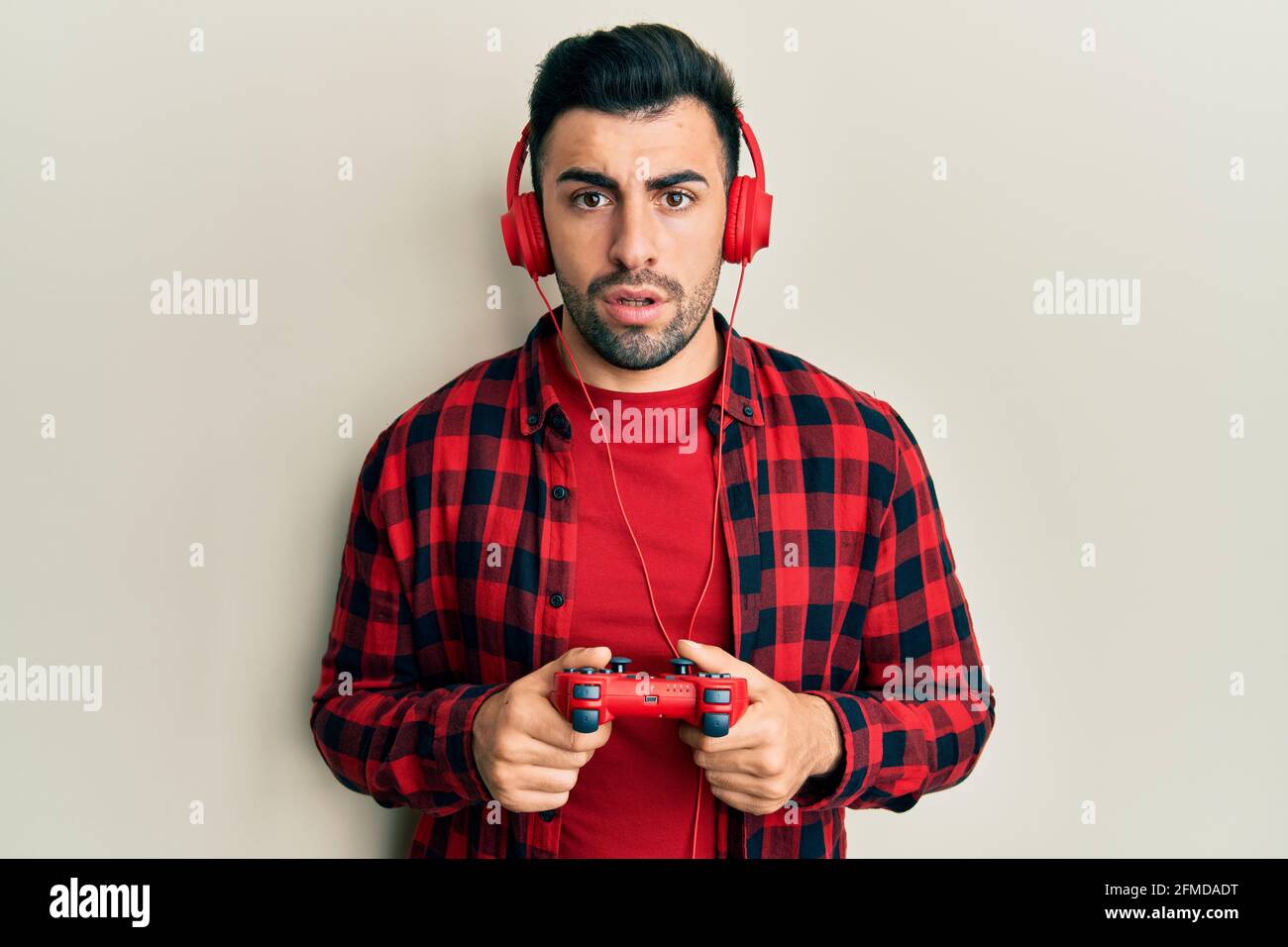 Young hispanic man playing video game holding controller in shock face ...