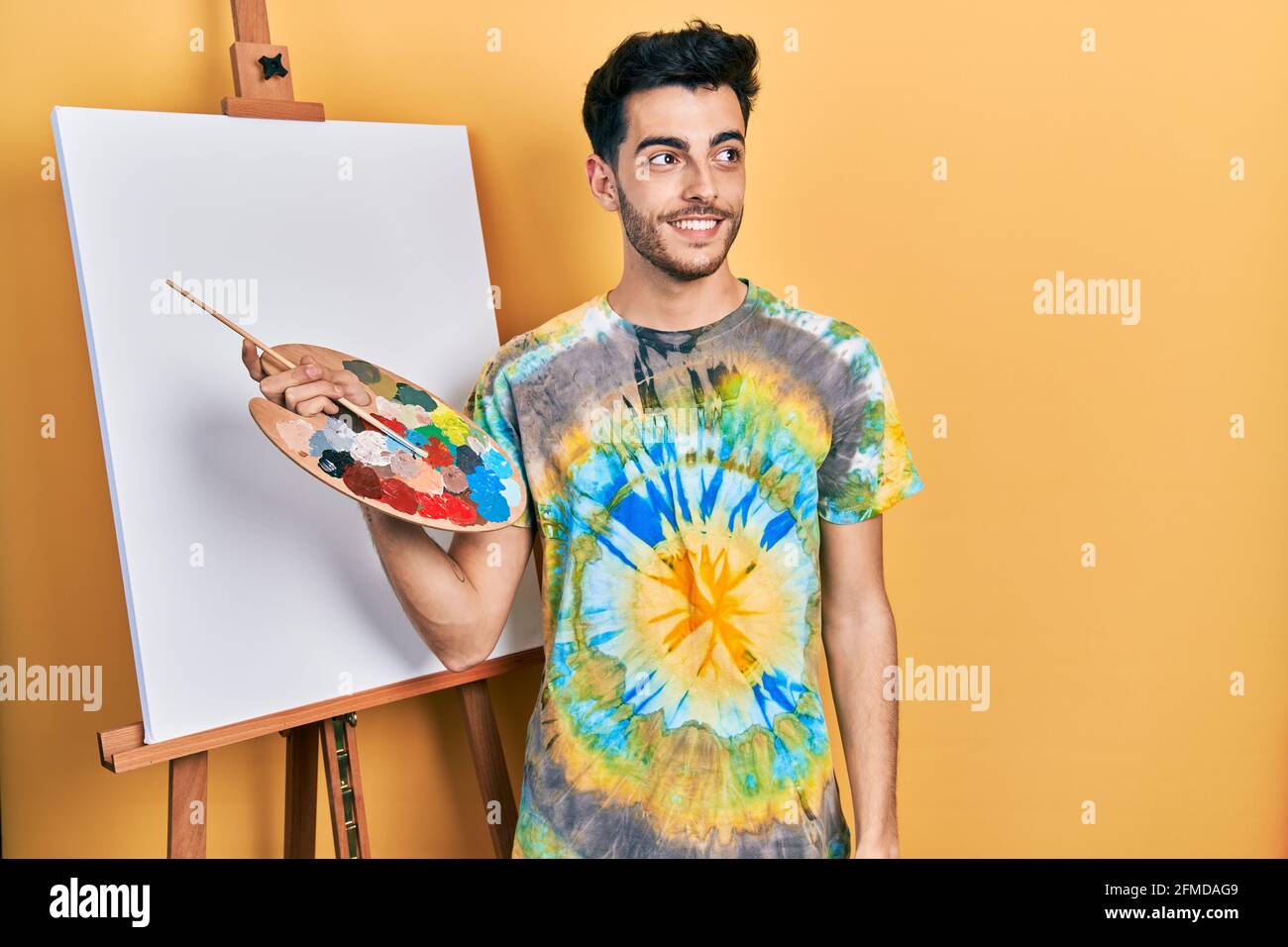 Young hispanic man standing drawing with palette by painter easel stand ...