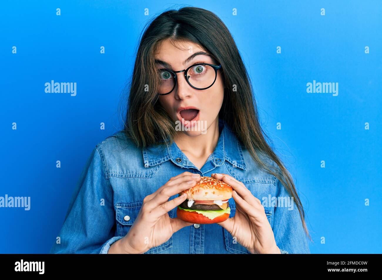 Young caucasian woman eating a tasty classic burger afraid and shocked ...