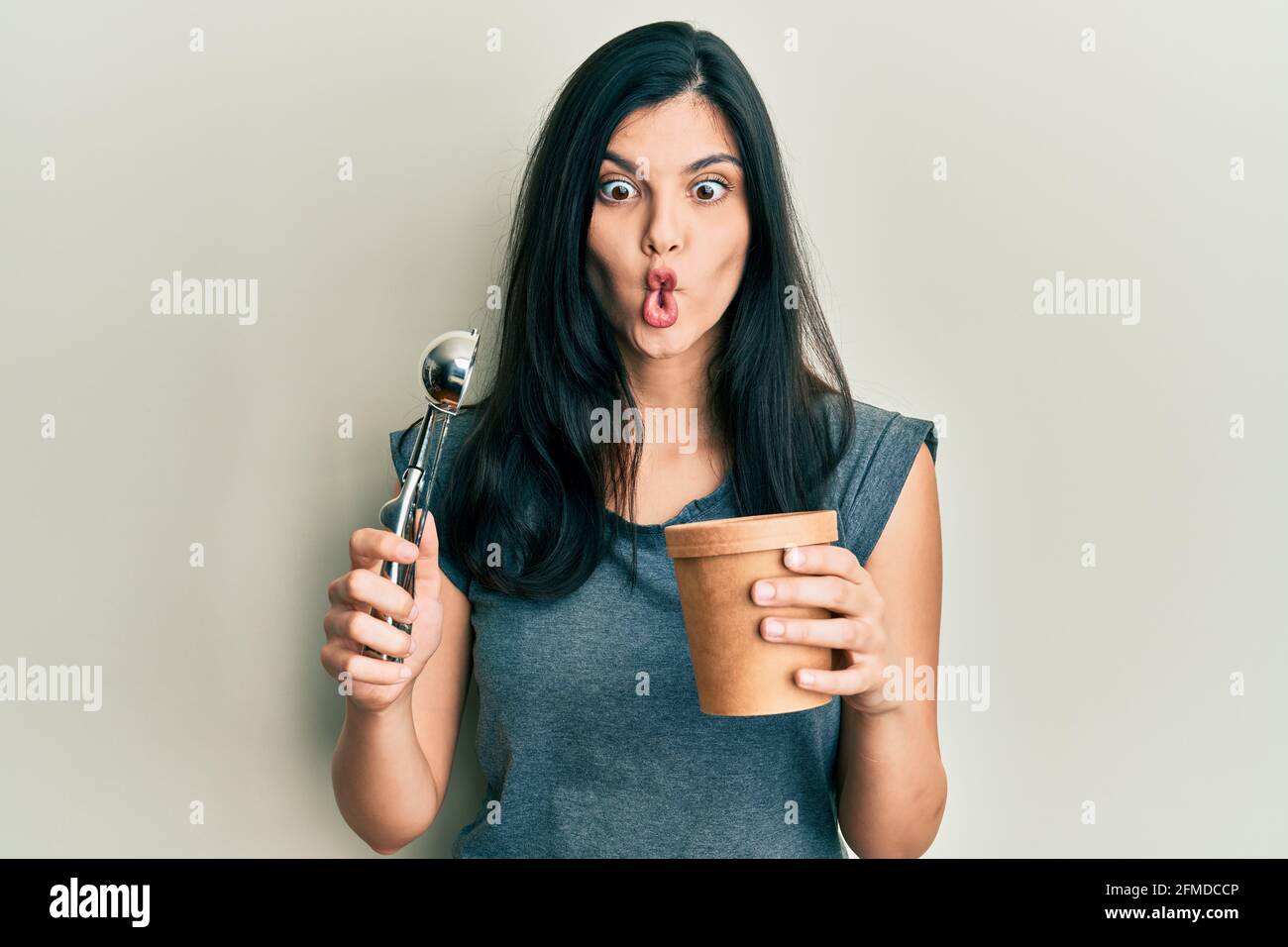 Young hispanic woman holding ice cream making fish face with mouth and ...