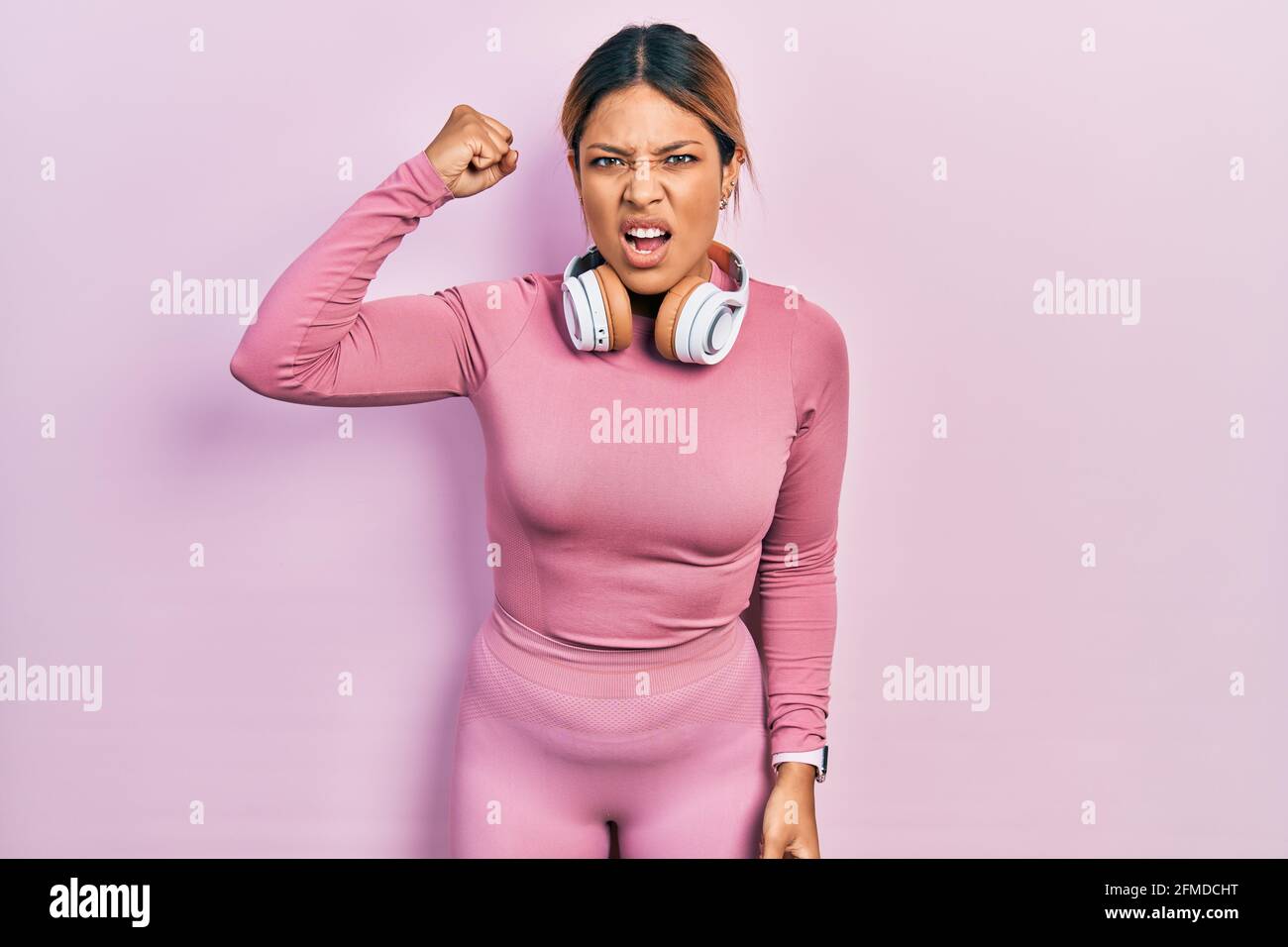 Beautiful hispanic woman wearing gym clothes and using headphones angry ...
