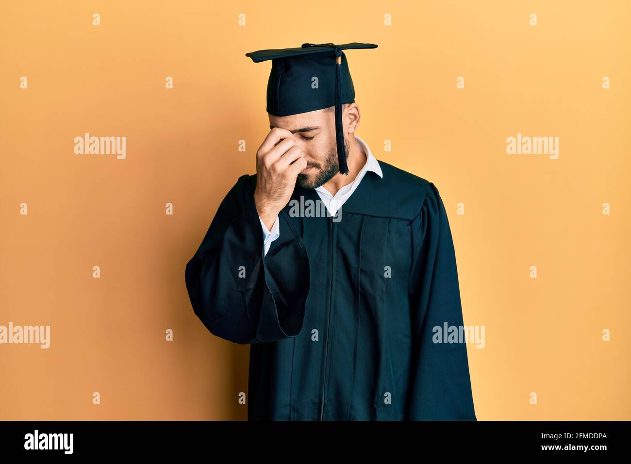 Young hispanic man wearing graduation cap and ceremony robe tired ...