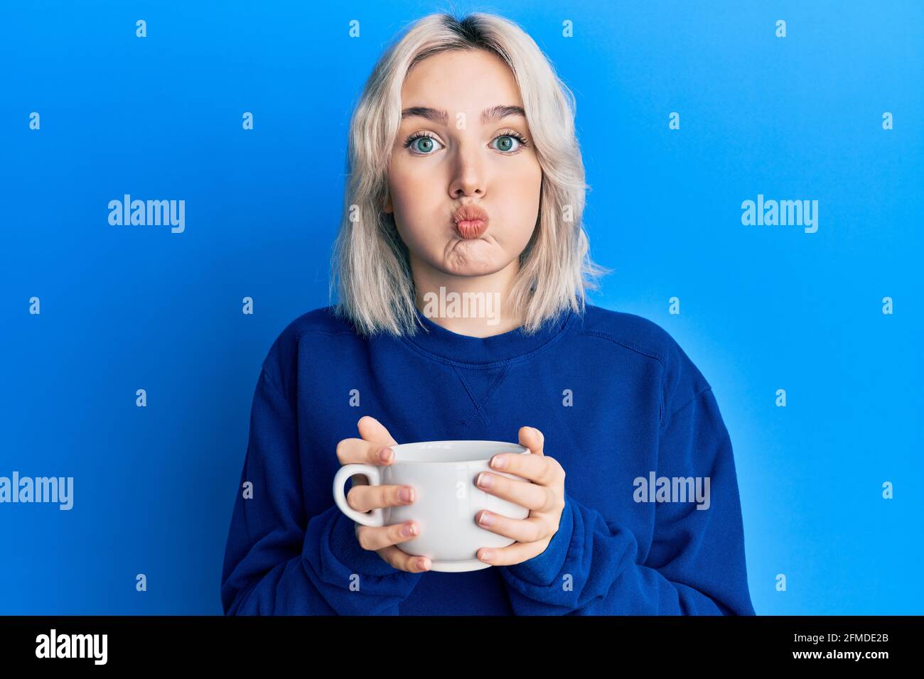 Young blonde girl drinking a cup of coffee puffing cheeks with funny ...